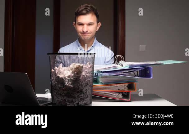 Stressed man at desk crumpling paper and tossing it into an overflowing ...
