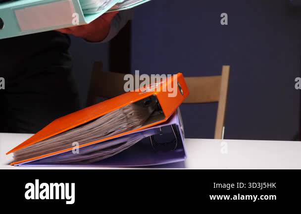 Person handles a towering stack of colorful ring binders overflowing with various documents ...