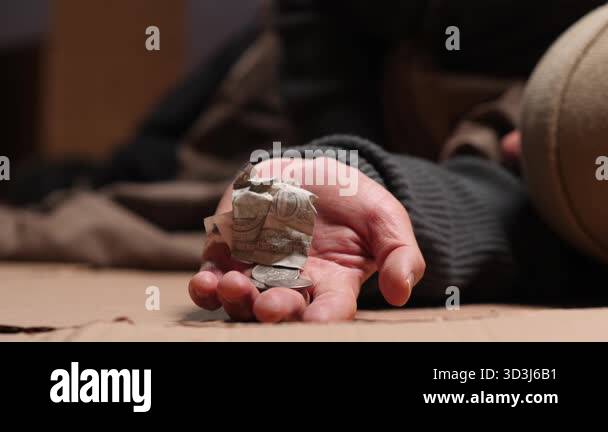 Homeless man lying on cardboard, his outstretched hand holding crumpled ...