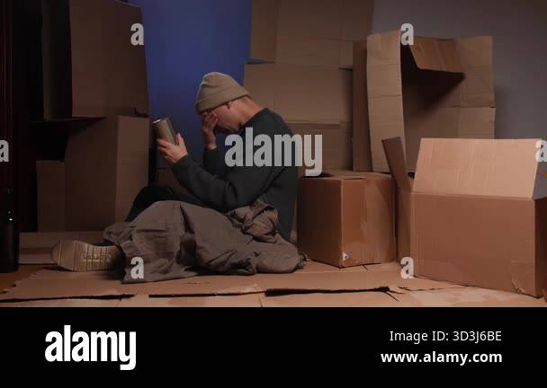 Homeless man sitting on cardboard boxes, holding a metal cup, covering ...