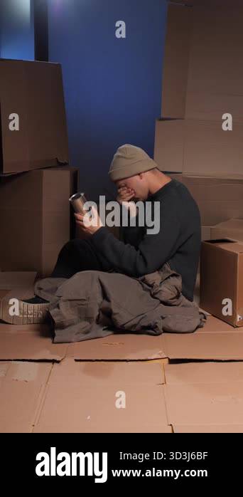 vertical video Homeless man sitting on cardboard boxes, holding a metal ...