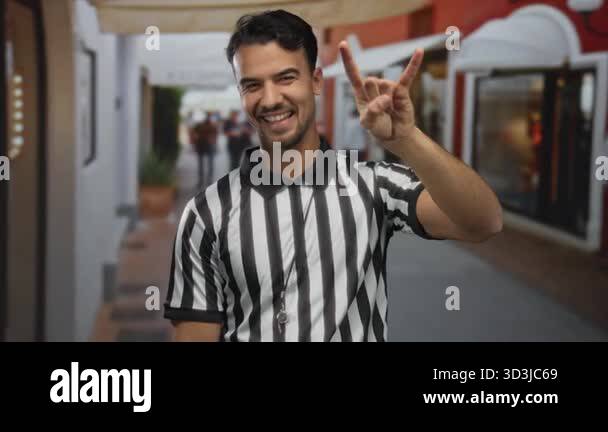 Young hispanic man in referee shirt smiling while making rock and roll ...