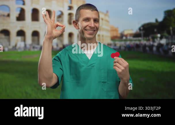 Doctor man in green uniform holds a small red heart and makes an okay ...