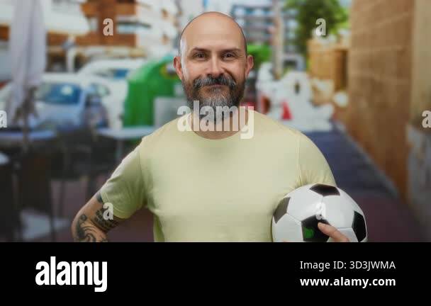 Bald man with beard holding soccer ball stands confidently on an urban ...