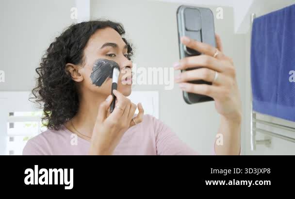 Non-binary adult using smartphone reflection applying black mask with wide makeup brush in ...