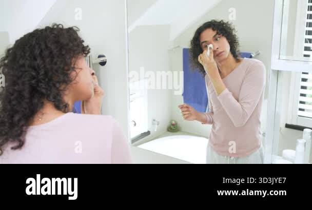 Non-binary adult glancing at mirror and wiping makeup with cotton pads ...
