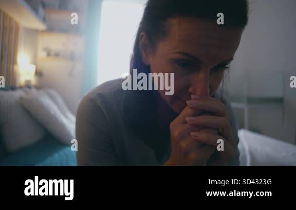 Woman sitting alone on bed, hands clasped and head lowered, expressing ...