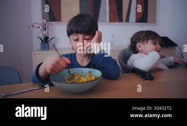 Young boy eating noodles with fork looking tired while baby sibling ...