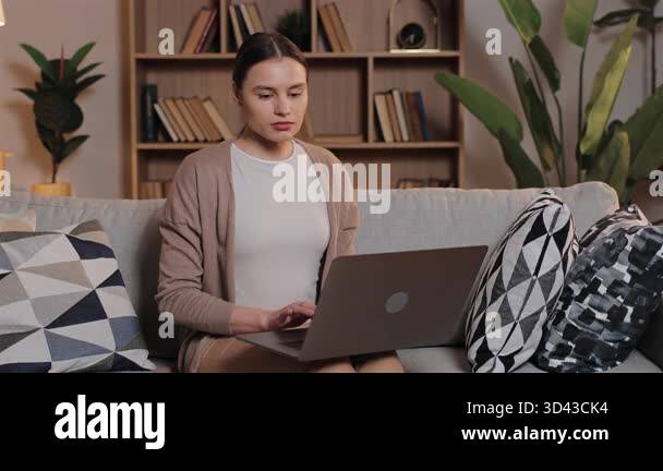 Focused Caucasian woman sitting on sofa using laptop. Looking at screen ...