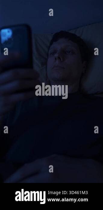 Tired man lies awake scrolling phone in dark bedroom. Blue screen light prevents sleep, showing ...