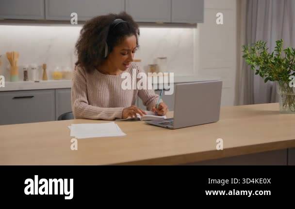 African American woman at home writing notes looking laptop listening ...