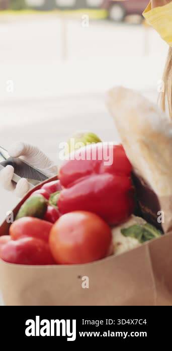 A woman wearing a yellow cap and face mask carries a paper bag filled ...