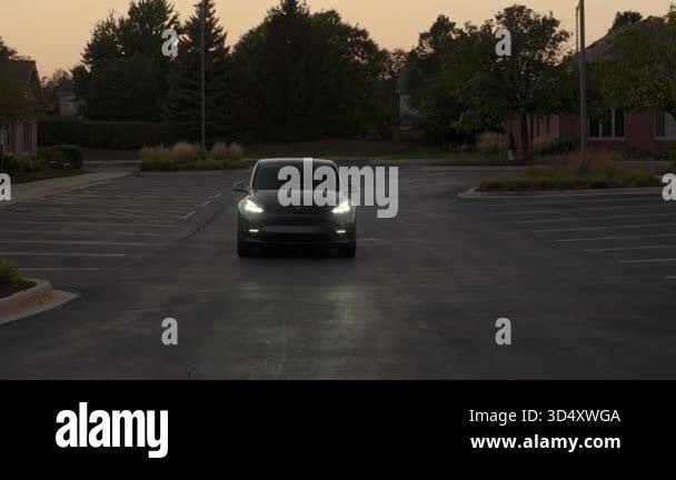Tesla drives through an suburban parking lot at dusk. The headlights ...