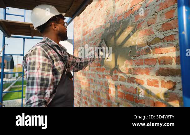 Diligent arab construction worker in a hard hat and overalls applying plaster to a brick wall ...
