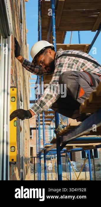 Diligent male builder wearing a hard hat and gloves using a spirit level to check the alignment ...