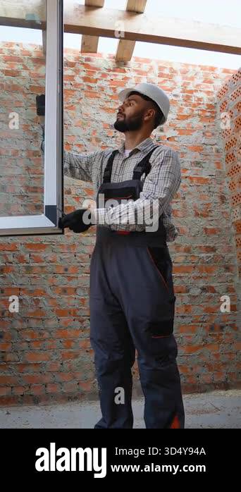 Diligent male builder wearing a hard hat and overalls carefully installing and adjusting a new ...