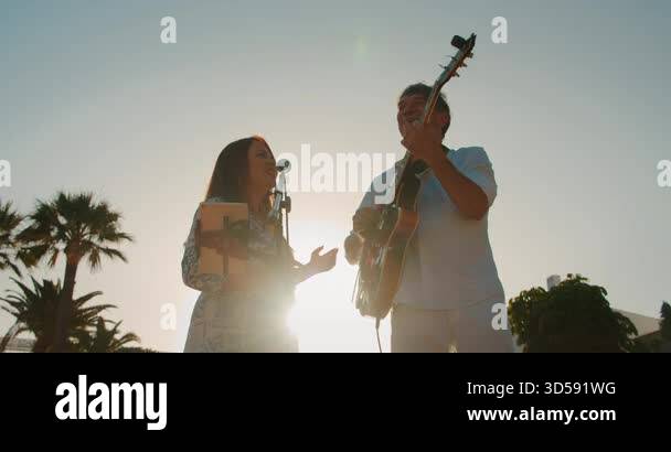 Singer and guitarist perform together under a clear sky with palm trees ...