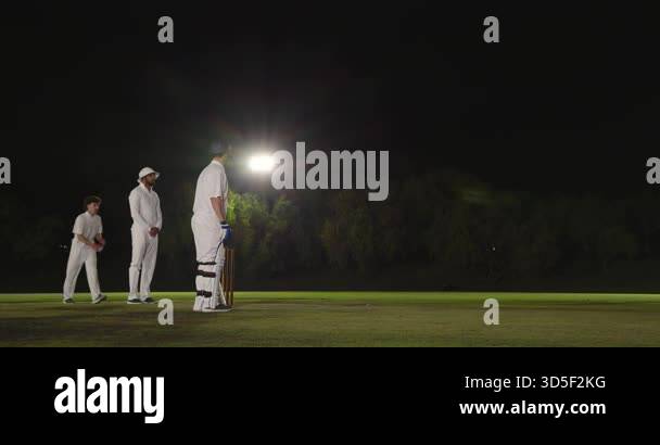 Running male cricketers on floodlit pitch delivering ball to batsman ...
