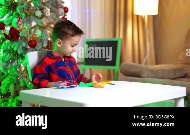 Beautiful baby boy in checkered shirt sits at desk. Kid takes scissors ...