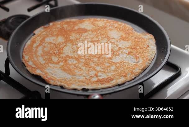 A golden-brown pancake cooking in a black frying pan on a gas stove ...