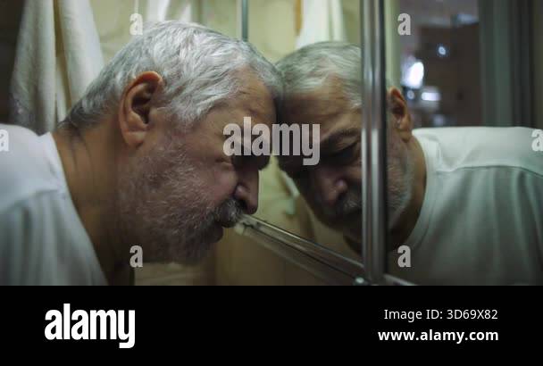 Older man resting his forehead against mirror, eyes closed, conveying ...
