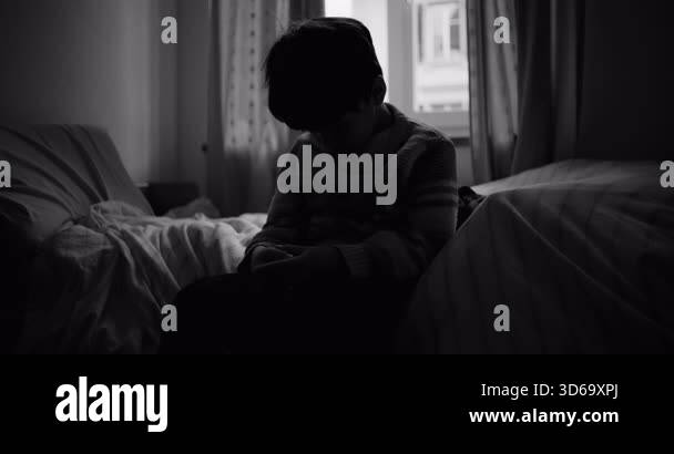 Boy sitting alone on bed with head bowed in dark bedroom near window ...