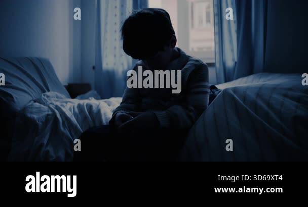 Child sitting on edge of bed in dim blue-lit room with head lowered ...
