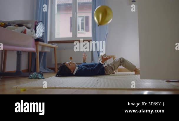 Child lying on bedroom floor tossing golden balloon into the air ...