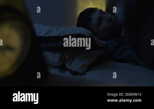 Tired man unable to sleep, looking at clock showing late night time ...