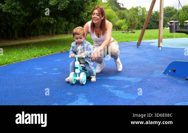 Happy mother assisting her toddler son learning to ride a balance bike on a wet playground ...