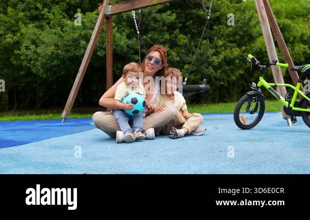 Happy family, a mother and her two sons are sitting on a playgrounds blue rubberized surface ...