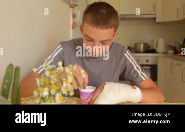 teen eating yogurt with arm cast at kitchen table, stirring cup with ...