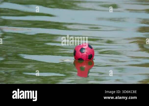 A small red soccer ball floats on a polluted pond with murky green ...