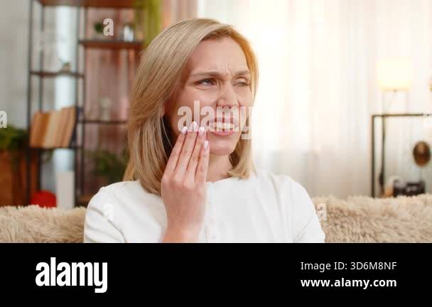 Middle-aged woman sits on home sofa holding cheek, wincing in visible ...