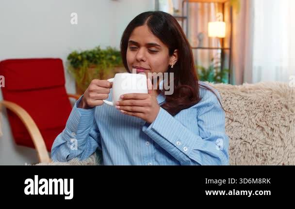 Indian young woman drinks warm tea on home sofa with relaxed expression ...