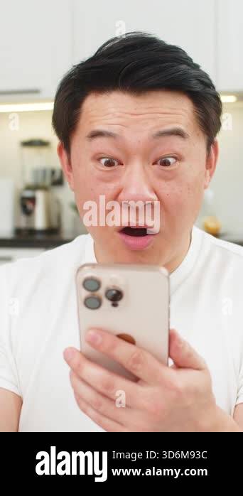 Asian man at home reads good news on smartphone says wow eyes wide smiles and starts typing ...