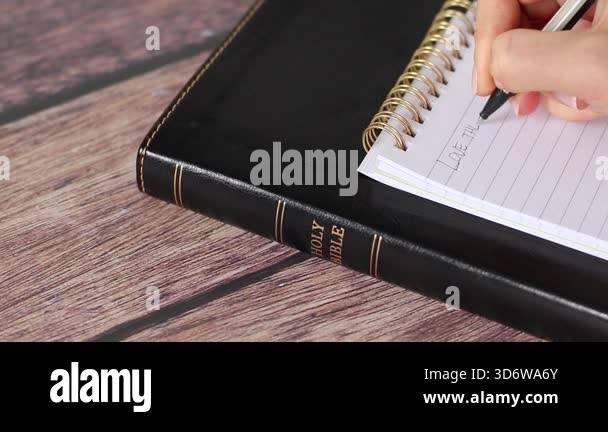 Hand is writing biblical quote to love God with all your heart in notebook on top of holy bible ...
