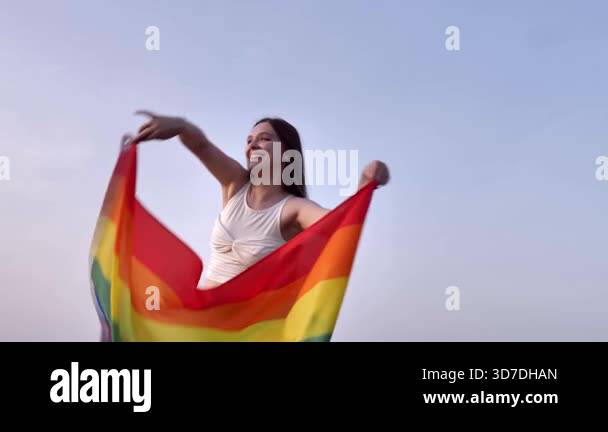 A smiling young woman holds the Rainbow Pride Flag above her head in a ...