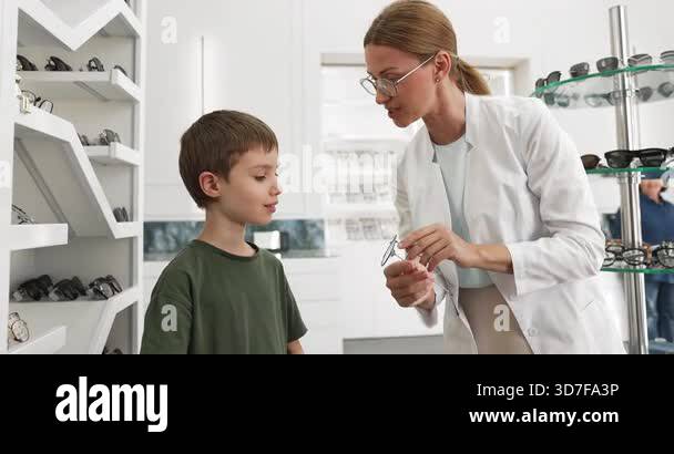 Friendly female optician in white coat helps to little adorable boy put ...