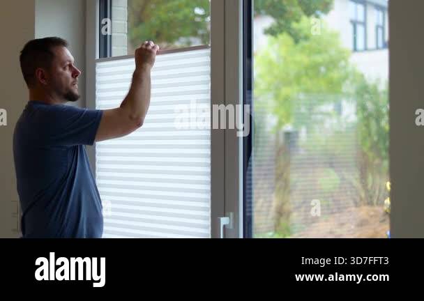 Close up of a caucasian man opening white blinds plisses on window ...
