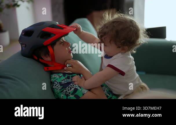 A toddler reaches up to touch and lift the older siblings helmet while ...