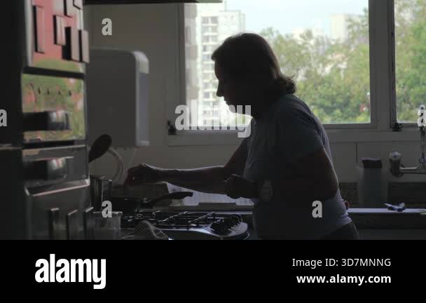 Elderly woman cooking alone in kitchen, silhouette by window conveying ...