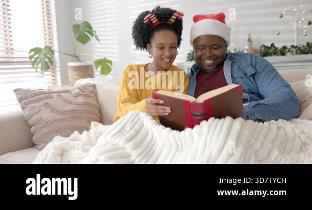 Diverse couple wearing Santa hat reading book aloud, turning pages ...