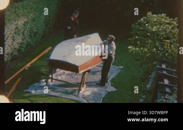 Two men work on a sailboat resting on sawhorses in a grassy backyard ...