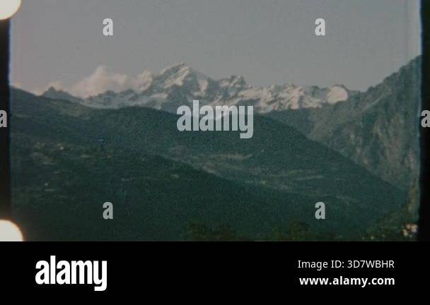 Retro 8mm film capturing a serene view of snow-capped mountain peaks against a clear sky. The ...