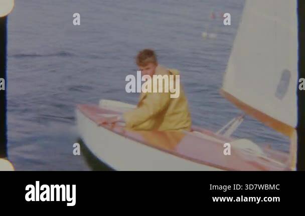 A man in a yellow raincoat sails a small boat on blue waters, captured ...