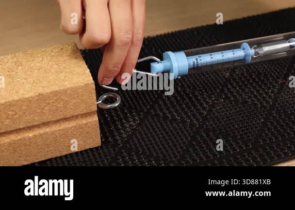 A hand conducts a friction test using a spring scale and wooden block ...