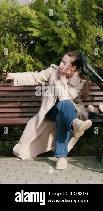 Female in beige coat captures a selfie while sitting on a bench next to ...
