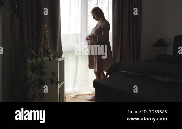 A young woman stands near the window in a softly lit room, gently ...