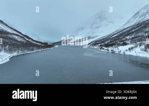A tranquil winter landscape features a calm alpine lake edged by snow ...